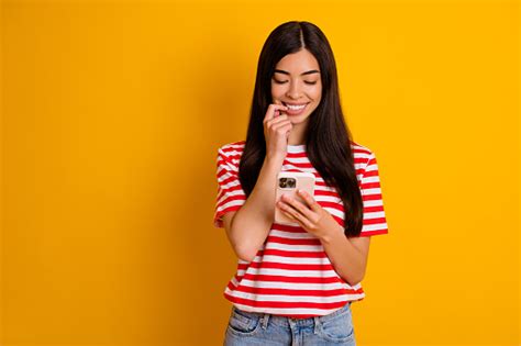 쾌활한 멋진 아가씨의 사진 물린 손가락 손톱 사용 전화 타이핑 블로그 게시물 착용 노란색 배경에 고립 된 빨간색 흰색 티셔츠 착용 매스컴에 대한 스톡 사진 및 기타 이미지