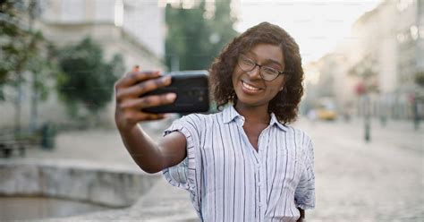 빨간 바지와 줄무늬 셔츠를 입고 야외에서 스마트폰으로 셀카를 찍고 고대 돌 분수 근처의 오래된 도시 거리에서 포즈를 취하는 쾌활한 젊은 아프리카계 미국인 사업가의 초상화