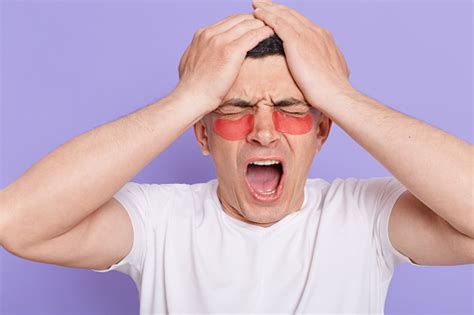 슬픈 분노의 감정을 표현하는 눈 아래에 보습 패치를 착용하고 큰 소리로 비명을 지르는 절망적 인 젊은 성인 남성은 머리와 눈을 감고 손을 얹습니다 35 39세에 대한 스톡