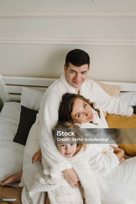 젊은 엄마 아버지는 침대에 누워 목욕 가운을 입은 귀여운 딸을 껴안고 호텔 방에서 카메라를 본다 의료 개념입니다 행복한 엄마 아빠 여자 아이가 웃고 있다 아이와 함께 즐거운