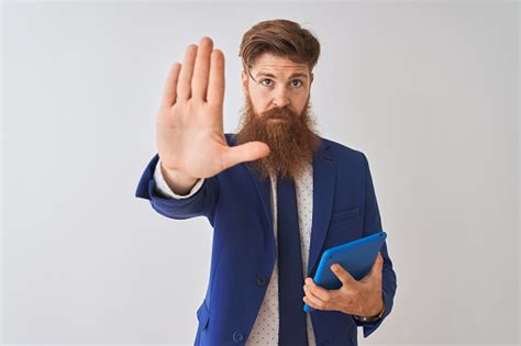 진지하고 자신감있는 표정 방어 제스처와 정지 기호를하고 열린 손으로 고립 된 흰색 배경 위에 태블릿을 사용하여 젊은 빨간 머리