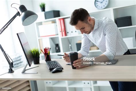 젊은 남자가 사무실에 테이블 근처 연필와 커피 한 잔을 들고 서 있다 젊은 사람이 문서와 컴퓨터와 함께 작동합니다 감독에 대한