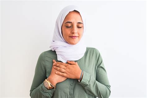 전통적인 이슬람 히잡을 입고 고립된 배경을 입은 젊은 아름다운 아랍 여성은 닫힌 눈과 얼굴에 감사하는 제스처로 가슴에 손을 대고 미소짓고 있습니다 건강 개념 몸짓에 대한