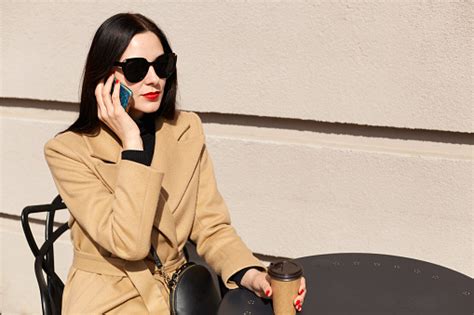 밝은 매력적인 젊은 여성은 전화를 통해 이야기 대화 그녀의 친구에 게 전화를 하 고 뜨거운 커피를 마시고 야외에서 빛 벽에 고립 된 앉아 평화로운 보인다 자유 시간