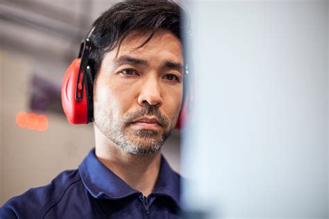 산업 시설 내부에서 귀 머프를 착용 한 중성인 아시아계 미국인 공장 노동자의 초상화를 클로즈업하십시오 귀 보호게에 대한 스톡