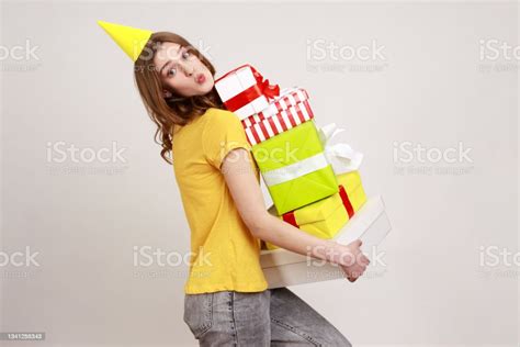 선물 상자를 많이 들고 머리에 갈색 머리와 파티 콘으로 매력적인 젊은 여성을 해치고 생일 선물을 포용 Pout 입술을 찾고