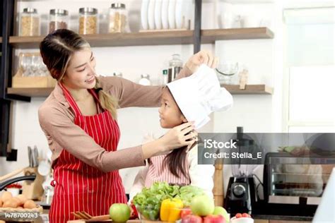 부엌에서 앞치마와 요리사 모자를 쓴 행복한 아시아 엄마와 딸 여자 아이를 위해 모자를 쓴 엄마 집에서 음식을 요리하는 가족 요리사 엄마와 아이가 함께 시간을 보내고 즐거운