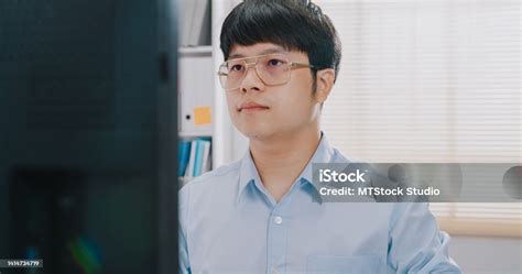 파란색 정장 셔츠를 입은 젊은 아시아 사업가는 책상 앞에 앉아 컴퓨터 타이핑을 살펴보고 소규모 사무실의 회의 게시판에서 프레젠테이션을 위해 서류 작업 마케팅을보고하는지