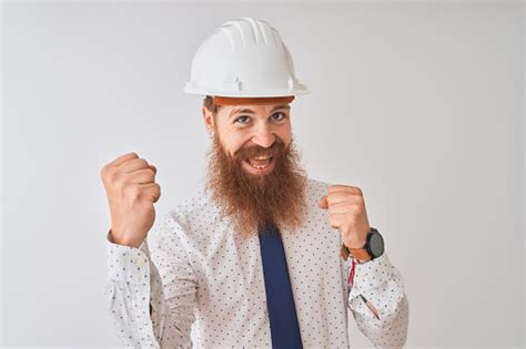 고립 된 흰색 배경 위에 보안 헬멧을 쓰고있는 젊은 빨간 머리 아일랜드 어 건축가 남자는 매우 행복하고 팔을 들고 미소를 지으며 성공을 위해 비명을 지르는 우승자 제스처를