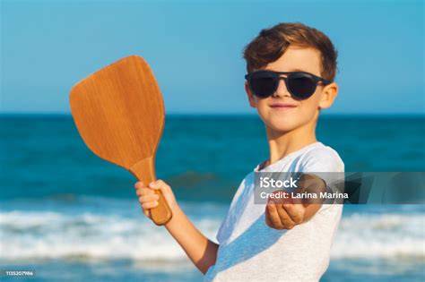 선글라스를 착용 한 소년은 비치 테니스를 합니다 아이는 그의 손으로 테니스와 공의 나무 테니스 라켓을 들고 푸른 하늘을 향하여 바다의 모래 해변에서 공을 봉사 할 준비를 하