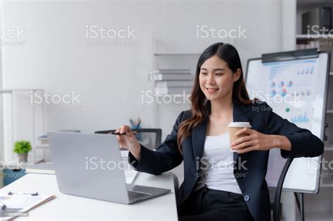 성공한 아시아 사업가가 노트북 컴퓨터를 사용하여 웃고 사무실에서 커피잔을 들고 있다 사무실에 행복하게 앉아 있는 자신감 넘치는 아시아 사업가 25 29세에 대한 스톡 사진