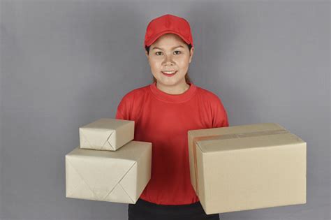 회색 배경에 소포 상자를 들고 서 있는 빨간 유니폼을 입은 행복한 배달 아시아 여성 30 34세에 대한 스톡 사진 및 기타 이미지 30 34세 개념 동남아시아 민족