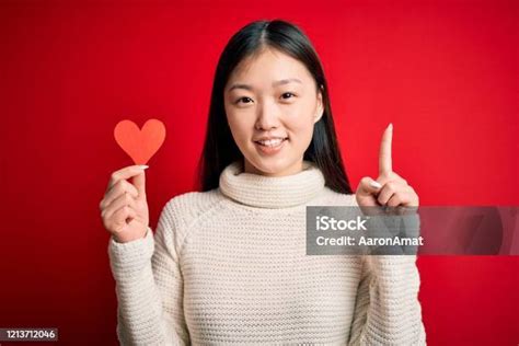 젊은 아시아 여자 들고 로맨틱 한 빨간 심장 종이 모양 위에 빨간 고립 된 배경 에 놀란 A 아이디어 또는 질문 가리키는 손가락 으로 행복한 얼굴 번호 하나 가리키기에 대한