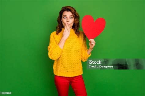 흥분 한 여자 보유 종이 하트 모양 엽서 숨기기 입 믿지 않는 잘 생긴 남자 초대 그녀의 날짜 착용 노란색 니트 스웨터 빨간 바지 고립 된 녹색 색상 배경 빨간 머리에 대한