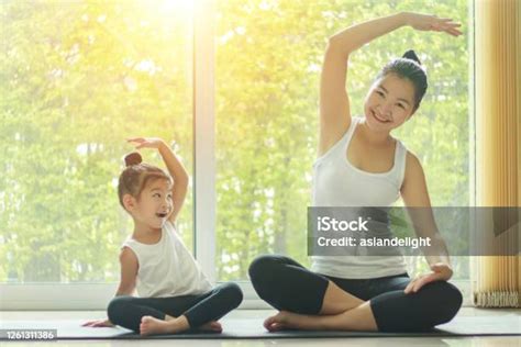 아시아 엄마는 집에서 운동하는 동안 웃는 얼굴로 어머니의 자세를 모방하려고 그녀의 옆에 앉아 사랑스러운 딸과 함께 집에서 요가를