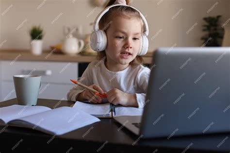 헤드폰을 끼고 웃고 있는 어린 소녀가 집에서 노트북을 사용하여 온라인으로 공부하고 이어폰을 낀 귀여운 행복한 어린 아이는 인터넷 웹 수업이나 Pc 수업을 받습니다