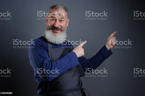 따라 오세요 여기를 봐 회색 배경에 고립 된 어깨 위에 빈 빈 장소를 가리키는 쾌활한 기쁘게 기뻐하는 숙련 된 앞치마를 입은 할아버지의 초상화를 닫습니다 교사 교육 관련