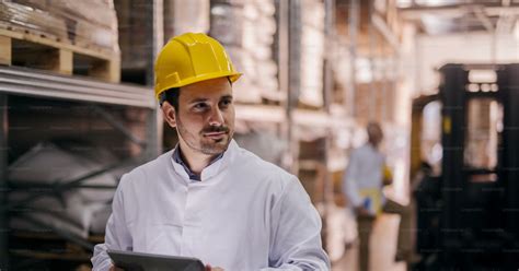 젊은 집중 남성 관리자가 공장에 서서 태블릿을 들고 있는 사진 머리에 헬멧을 쓴 흰 코트를 입고 만족스럽고 자랑스러워 보입니다 사진 Unsplash의 네덜란드 이미지