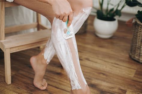 탈모 크림 개념으로 제모 흰 수건을 쓴 젊은 여성이 다리에 면도 크림을 바르고 녹색 식물이 있는 집 욕실에서 플라스틱 면도기를 들고 있다 스킨 케어 사진