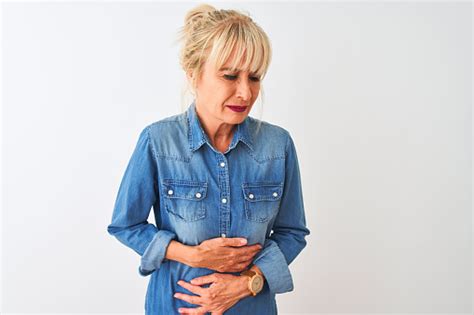 캐주얼 한 데님 셔츠를 입고 있는 중년 여성은 위장에 손을 얹고 고립된 흰색 배경 위에 서 있기 때문에 메스꺼움 고통스러운 질병이 기분이 나아감에 좋다 아쉬 컨셉 한 명의