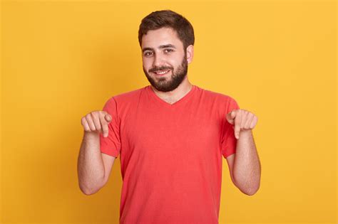 카메라를 직접 바라보며 캐주얼한 밝은 빨간 티셔츠를 입고 흰색 스튜디오 벽에 서서 손가락을 아래로 가리키는 젊은 남성의 수평