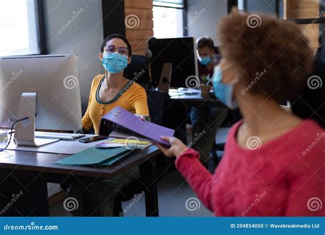 Diverse Female Colleagues Wearing Face Masks Passing File Between Desks