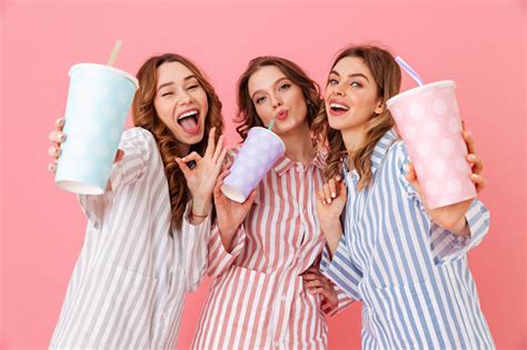 핑크 배경에 고립 된 재미있는 파자마 파티 동안 종이 컵에서 미소와 차가운 탄산음료를 마시는 다채로운 줄무늬 잠옷에 좋은 분위기를 가진 세 가지 콘텐츠 여성 3 명에 대한