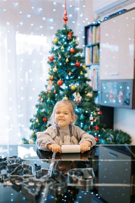 Little Smiling Girl With A Small Rolling Pin Sitting At A Table Near A Decorated Christmas Tree
