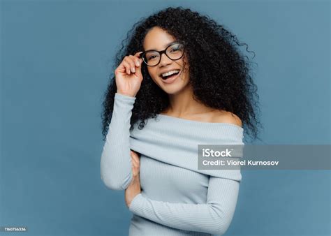 미소 젊은 아프리카 계 미국인 여성의 고립 된 샷은 안경의 프레임을 터치 기쁜 표정을 가지고 파란색 배경에 고립 된 세련된 점퍼를 입고 친구와 함께 자유 시간을 보내고 감정