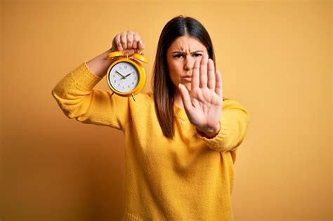 진지하고 자신감있는 표정 방어 제스처로 정지 표지판을하는 열린 손으로 고립 된 노란색 배경 위에 서있는 알람 시계를 들고있는 젊은 아름다운 여성 경계 표지에 대한 스톡 사진