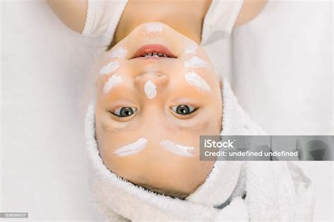 그녀의 뺨 코와 이마에 얼굴 크림 고립 된 흰색 배경에 누워 흰색 목욕 수건에 싸여 작은 귀여운 두 살 짜리 아기 소녀의