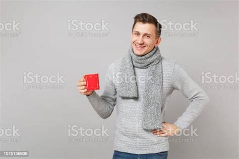 쾌활 한 젊은 남자가 회색 스웨터 스카프 커피 또는 차 스튜디오에 회색 배경에 고립의 빨간 컵을 들고 건강 한 생활 습관 병