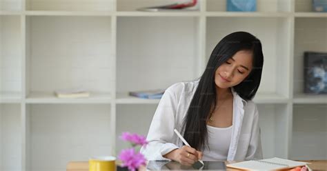 흰색 셔츠를 입은 젊은 아름다운 여성이 스타일러스 펜과 컴퓨터 태블릿으로 작업하면서 거실 책장 위의 나무 작업 테이블에 배경으로