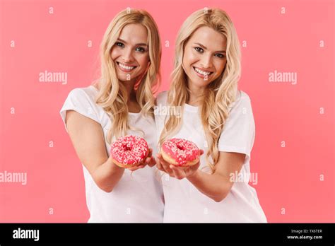 Smiling Blonde Twins Wearing In T Shirts Giving Donuts At The Camera Over Pink Background Stock