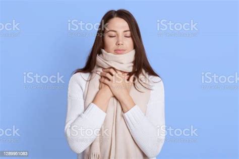 흰색 캐주얼 셔츠를 입은 젊은 여성이 스카프에 싸여 목이 아환하고 목에 손을 얹고 화가 났습니다 인후통 고통스러운 삼키기 푸른 배경 위에 고립 된 포즈를 취하는 어두운 머리