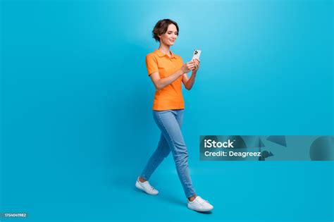 좋은 여자의 전체 길이 사진 V 기호를 보여주는 오렌지 티셔츠를 입고 스마트 폰을 보면 파란색 배경에 고립 된 빈 공간으로 이동합니다 여자에 대한 스톡 사진 및 기타 이미지