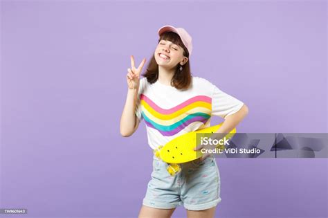보라색 파스텔 배경에 고립 눈을 폐쇄 유지 승리 기호를 보여주는 노란 스케이트 보드와 생생한 옷에 웃는 십 대 소녀 사람들은 진심으로 감정 라이프 스타일 개념입니다 복사