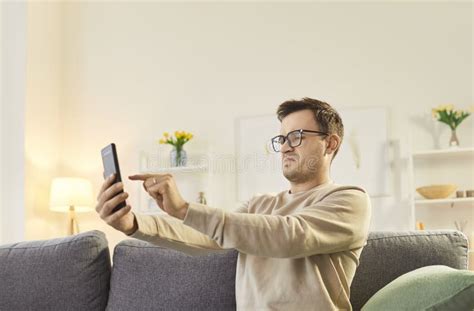 Young Man In Glasses With Vision Problems Holding Smartphone At Arm Length Squinting At Screen