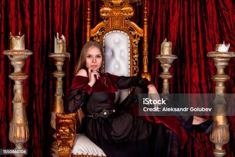 골동품 왕좌 요새에 앉아 카메라를 바라보는 역사적인 이미지의 귀여운 아가씨 옛 스타일의 중세 여왕이 성의 방에서 황금 왕좌에 포즈를 취하고 있다 극장 공연의 개념입니다