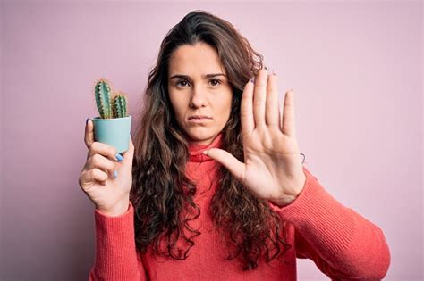 진지하고 자신감있는 표정으로 정지 표지판을하고 열린 손으로 고립 된 분홍색 배경 위에 작은 선인장을 들고 곱슬 머리를 가진 젊은 아름다운 여자 방어 제스처 경계 표지에 대한