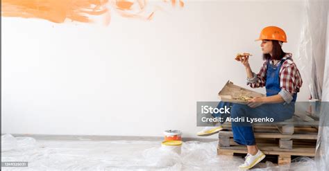 여성 수리인이 페인트로 벽을 그렸고 수리를 하고 맛있는 피자를 먹는 나무 팔레트에 앉아 있습니다 오렌지 색 하드 모자를 쓴 소녀