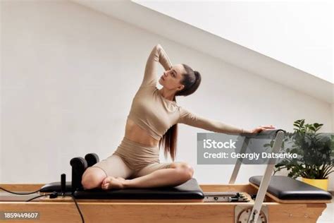 한 어린 소녀가 밝은 스튜디오의 리포머 침대에서 필라테스를 하고 있다 베이지색 보디수트를 입은 아름다운 날씬한 갈색 머리는