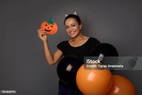 고양이 귀와 후프를 입고 검은 색 옷을 입고 화려한 검은 색과 오렌지 풍선을 들고 펠트 컷 수제 호박을 보여주고 카메라를보고 미소 이빨 미소를 짓는 젊은 예쁜 여자 가을에