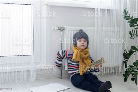 아이는 스웨터와 모자를 따뜻하게 입고 라디에이터에 앉아 있습니다 경제 위기의 개념과 가정의 열과 난방 부족 2 3 살에 대한 스톡 사진 및 기타 이미지 Istock