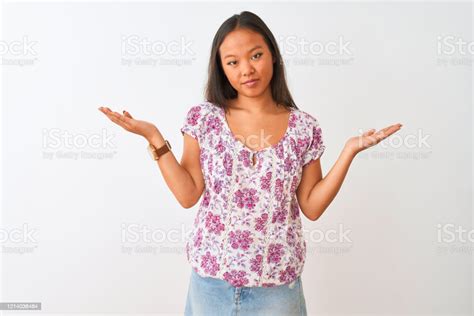 고립된 흰색 배경 위에 서있는 캐주얼 한 꽃 무늬 티셔츠를 입은 젊은 중국 여성은 팔과 손을 들고 혼란스러운 표정을 짓고 있습니다 의심 개념 T 셔츠에 대한 스톡 사진 및