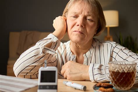 나이 지긋한 여성이 탁자에 앉아 턱을 괴고 혈당계를 유심히 바라보고 있다 그녀 옆에는 차 한 잔과 쿠키 한 접시가 놓여 있어 건강에 대한 성찰의 순간을 보여줍니다 55 59