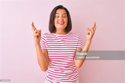 줄무늬 티셔츠를 입은 젊은 아름다운 여성은 고립 된 분홍색 배경 제스처 손가락 위에 서서 희망과 눈을 감고 미소를 지었습니다 행운과 미신 개념 T 셔츠에 대한 스톡 사진 및