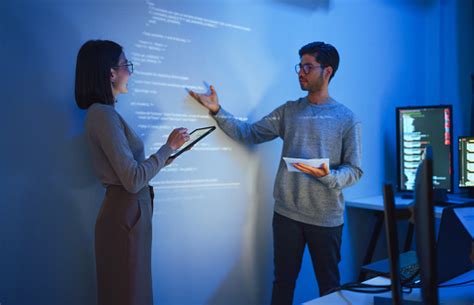 아시아 출신의 두 소프트웨어 개발자가 협업 코딩 세션에서 코드를 벽에 투사하고 여러 컴퓨터 화면을 표시하며 프레젠테이션을 합니다