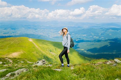 산을 배경으로 산 능선의 경사면에 서 있는 행복한 금발 여성 등산객의 초상화 배경에 푸른 흐린 하늘 여자는 행복하고 카메라를보고 있습니다 여행과 활동적인 라이프 스타일 개념