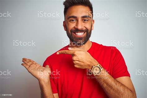 고립 된 흰색 배경 위에 빨간 티셔츠를 입은 젊은 인도 남자는 손으로 손가락으로 가리키는 동안 카메라에 놀랍고 미소를 지었습니다 남자에 대한 스톡 사진 및 기타 이미지